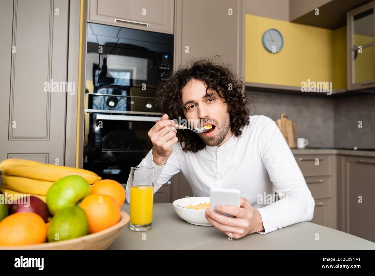 handsome young man eating flakes and looking at camera while using ...