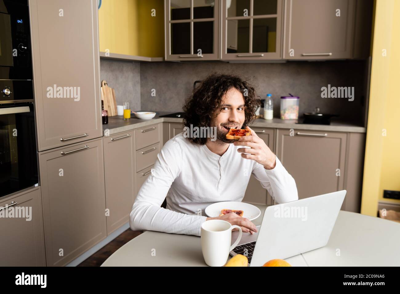 cheerful freelancer eating toast and looking at camera near laptop and ...