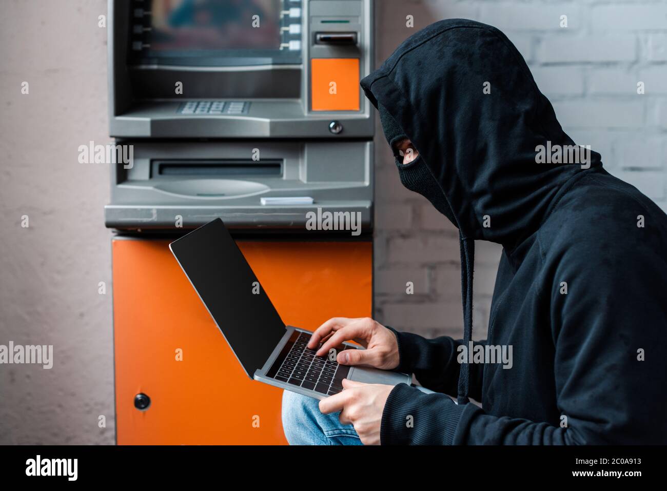 Side view of hacker in mask using laptop with blank screen near atm ...
