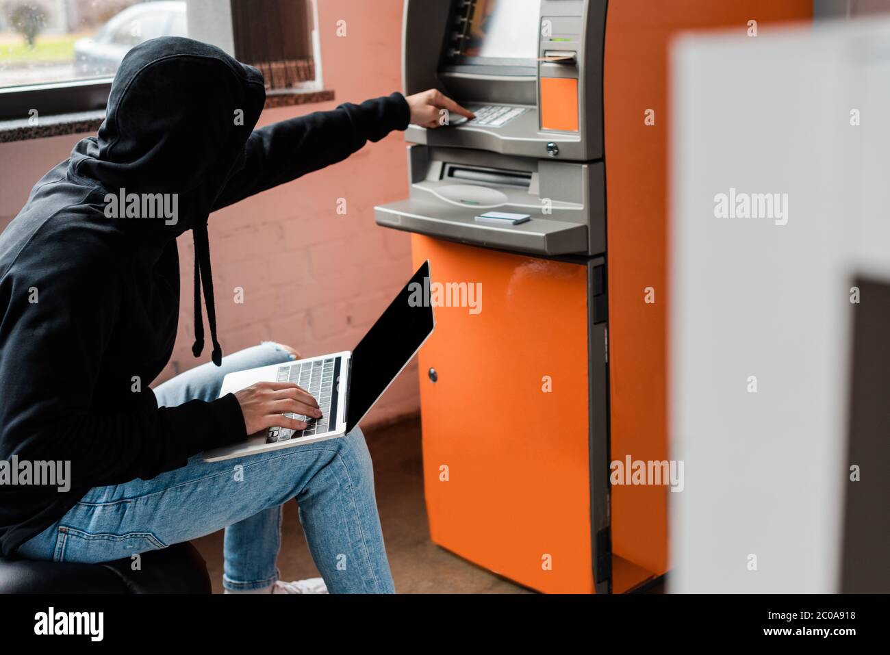 Side view of hacker in mask using atm and laptop with blank screen ...