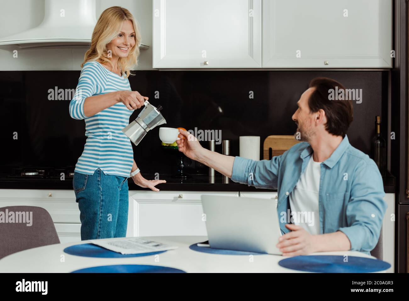Selective focus of smiling woman pouring coffee near husband and laptop ...