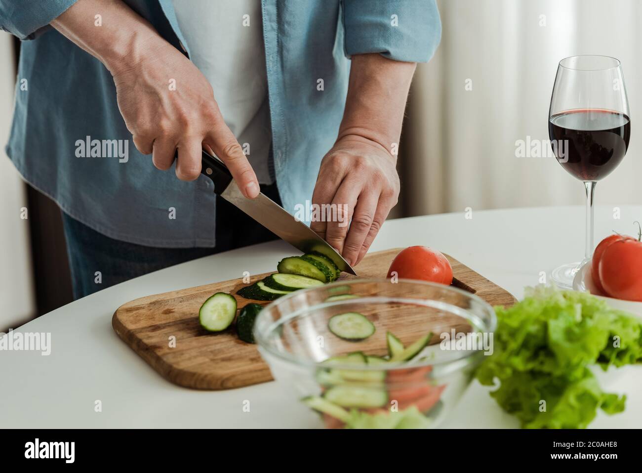 Cropped view of man cutting cucumber while cooking salad near glass of ...