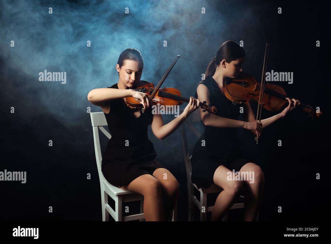 attractive female musicians playing on violins on dark stage with smoke ...