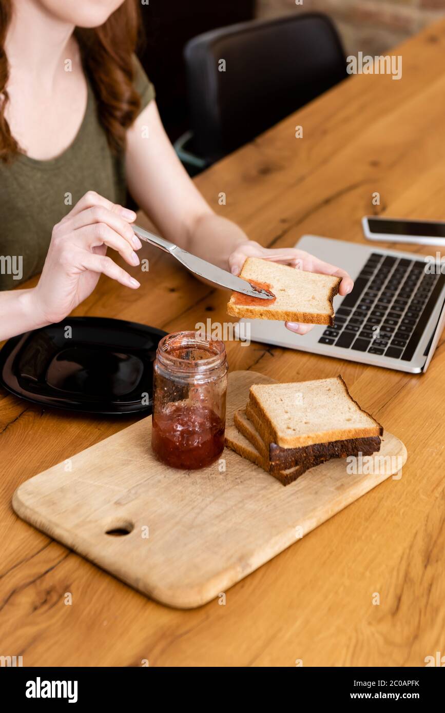 Cropped view of woman pouring jam on toast near gadgets on wooden table ...