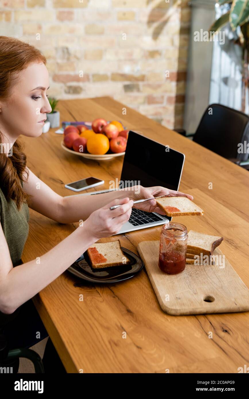 Side view of woman pouring jam on toast near digital devices on table ...