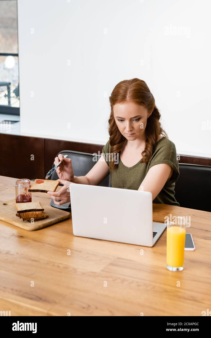 Selective focus of woman looking at laptop while holding toast with jam ...