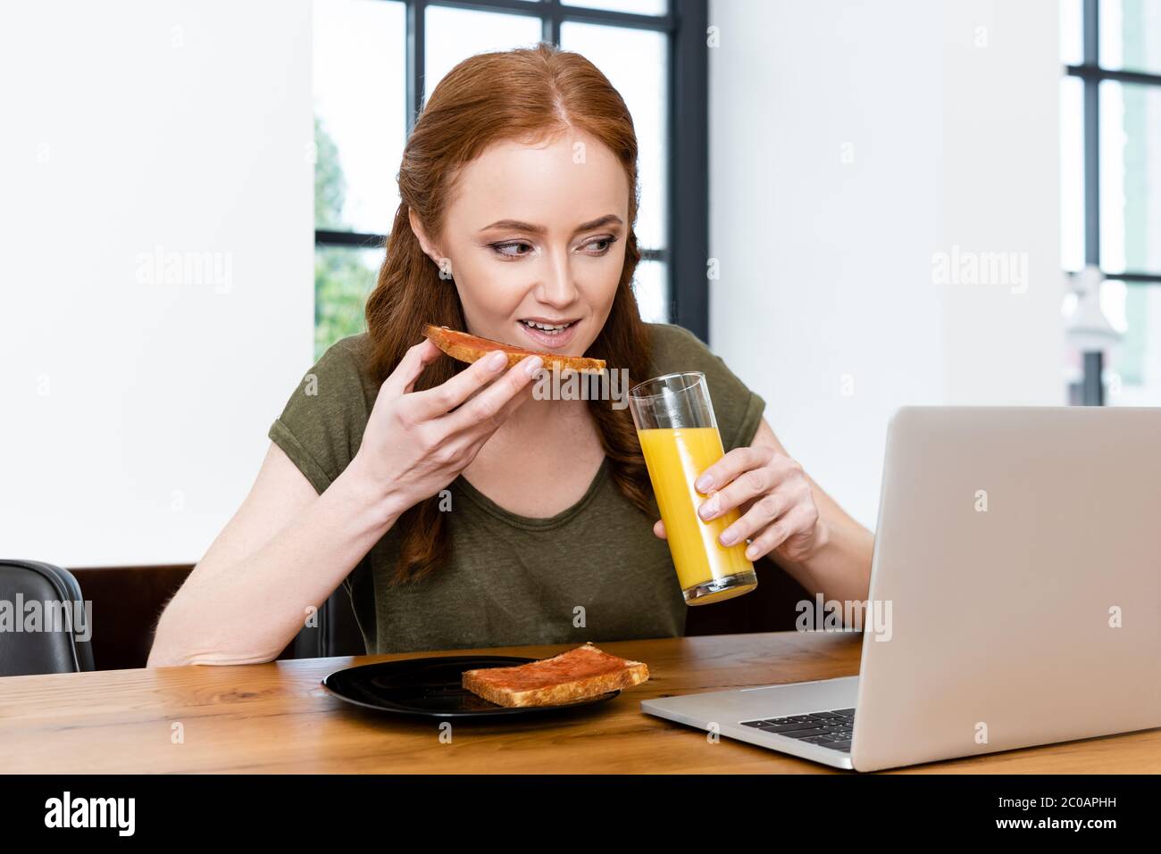Woman eating toast and holding glass of orange juice near laptop on ...