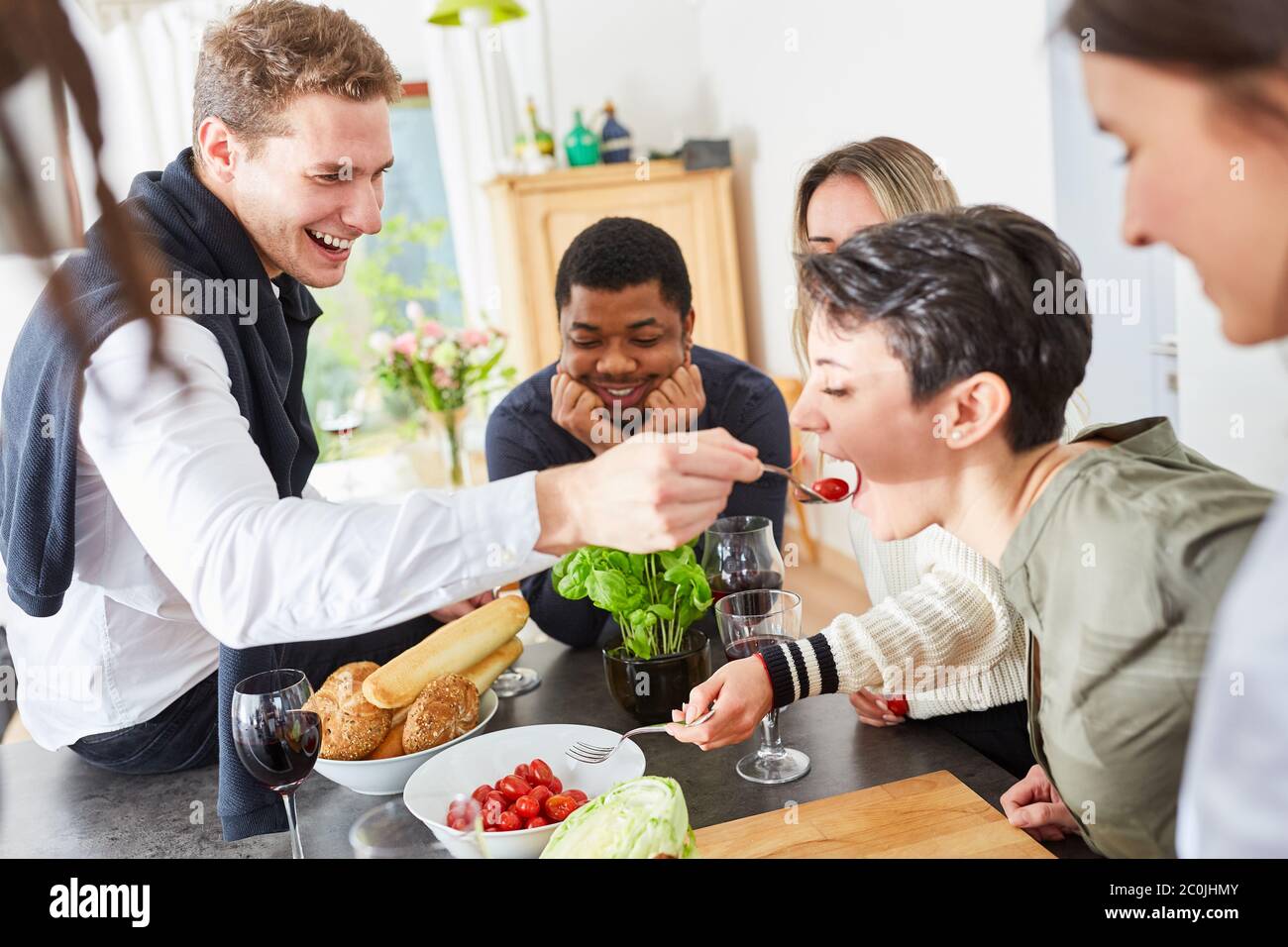 Laughing friends in the kitchen prepare food and taste a red tomato ...
