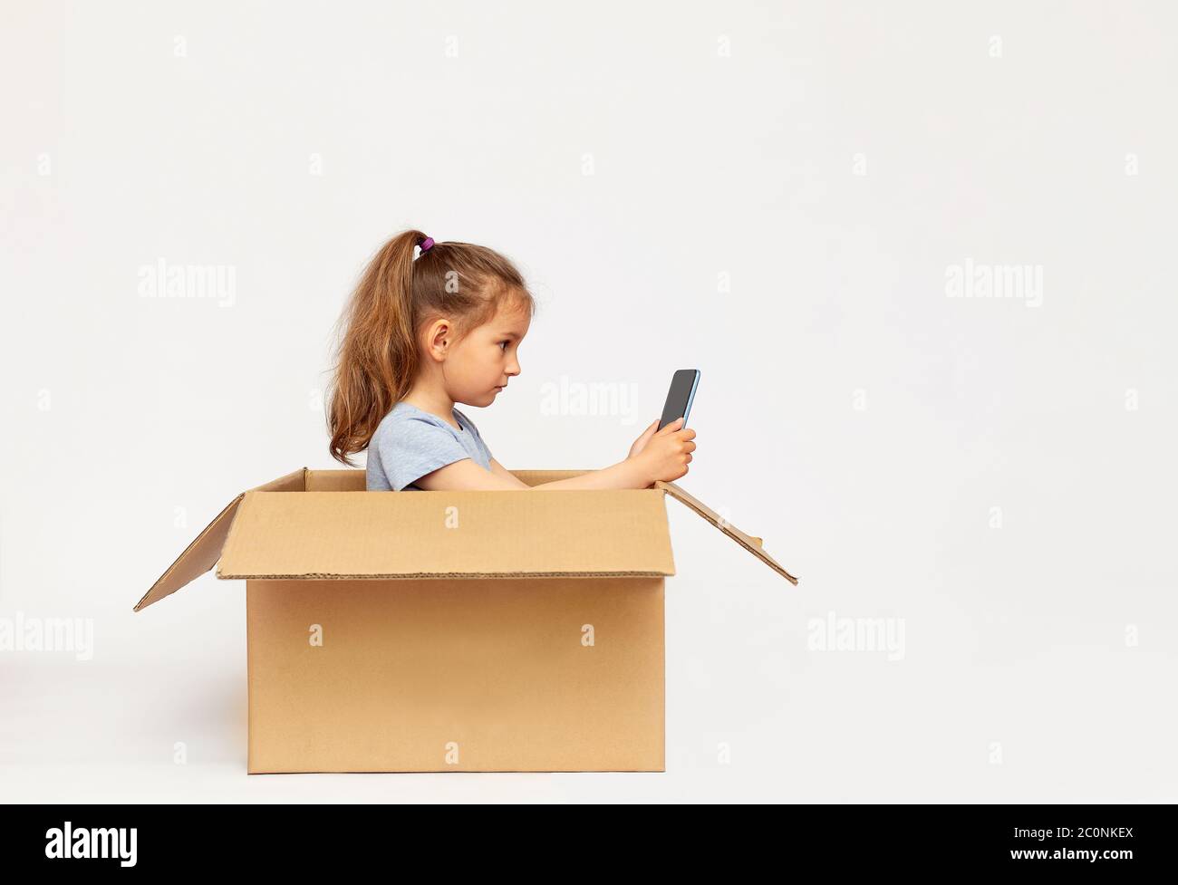 A little girl sits in a large cardboard box and plays on the tablet ...