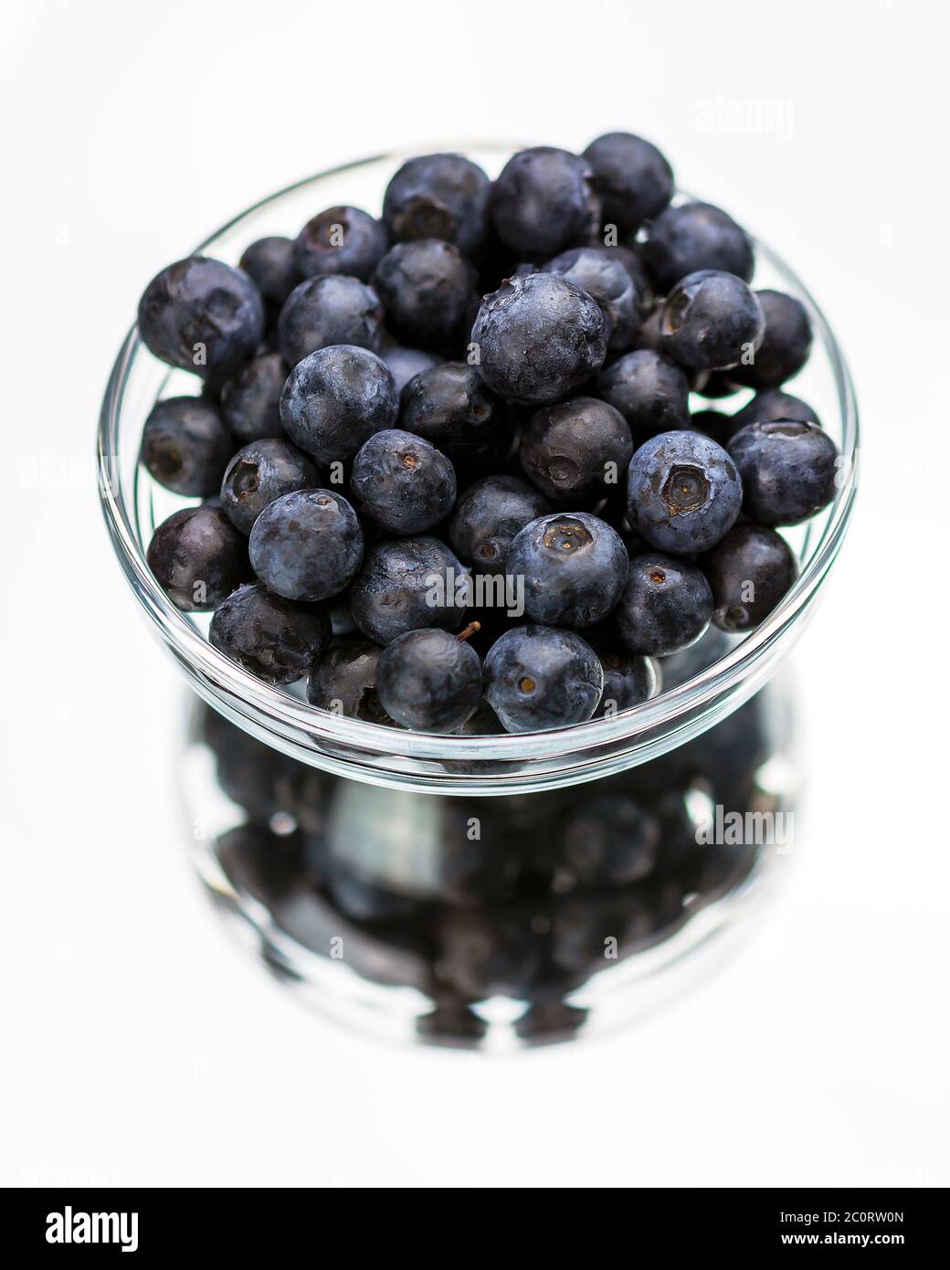 A clear glass bowl of blueberries on a mirror with reflection. White ...