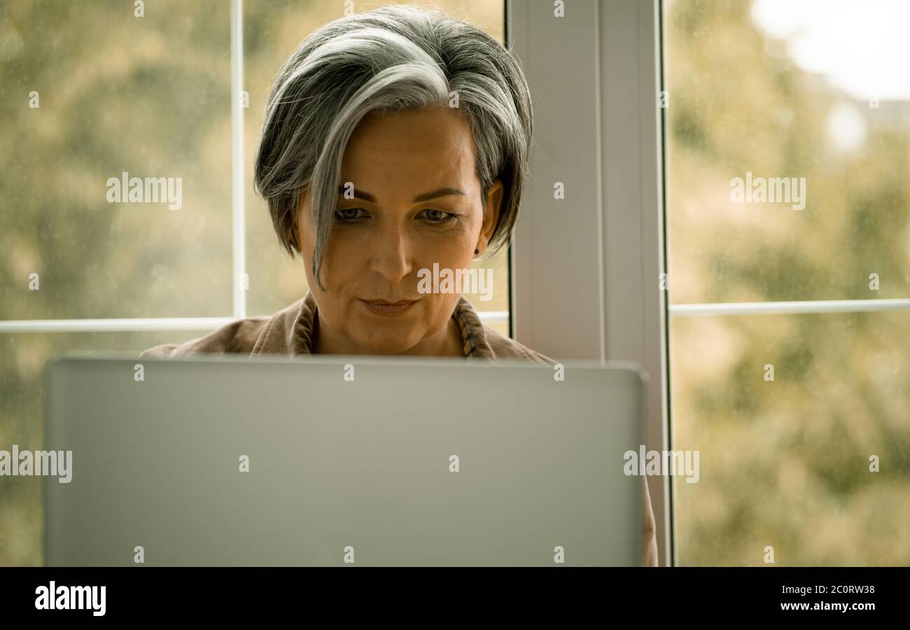 Mature woman works laptop sitting on sill. Serious Caucasian Lady ...