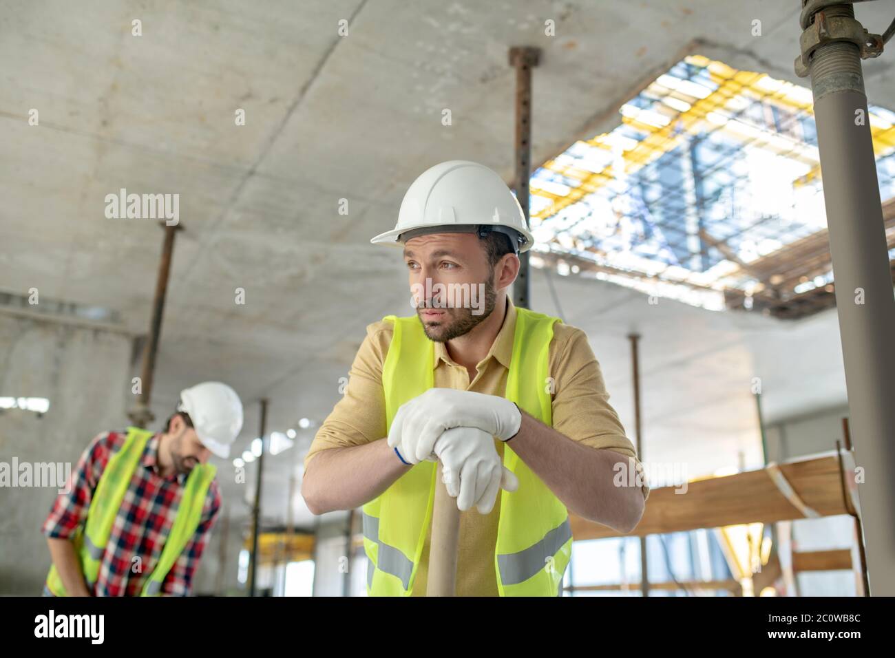 Tired building worker in yellow vest and gloves leaning on handle ...