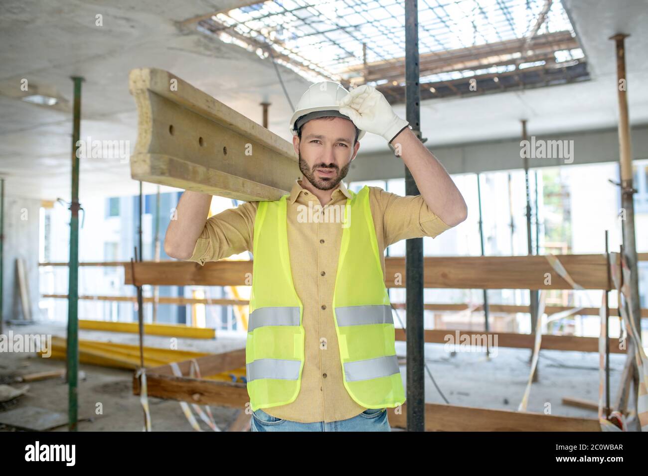 Tired building worker in yellow vest and gloves carrying wooden board ...