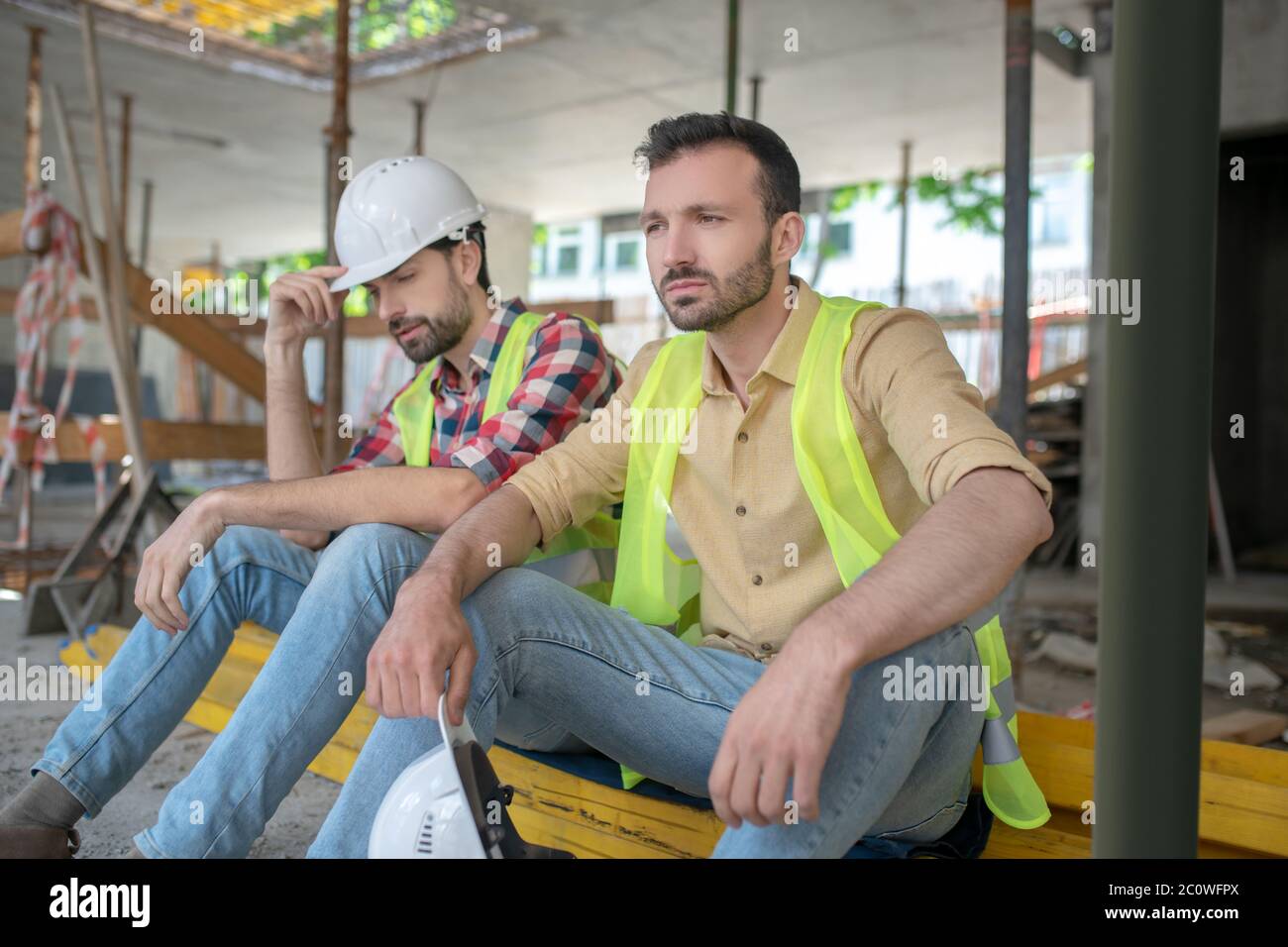 Tired building workers in yellow vests sitting on boards, one of men ...