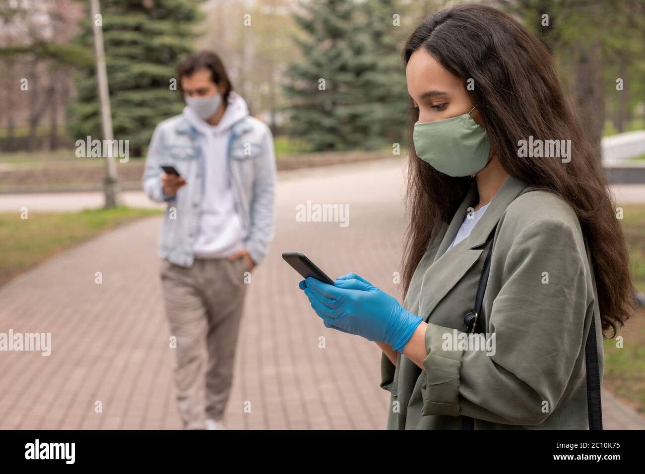 Young woman in green mask and gloves standing in city park and texting ...
