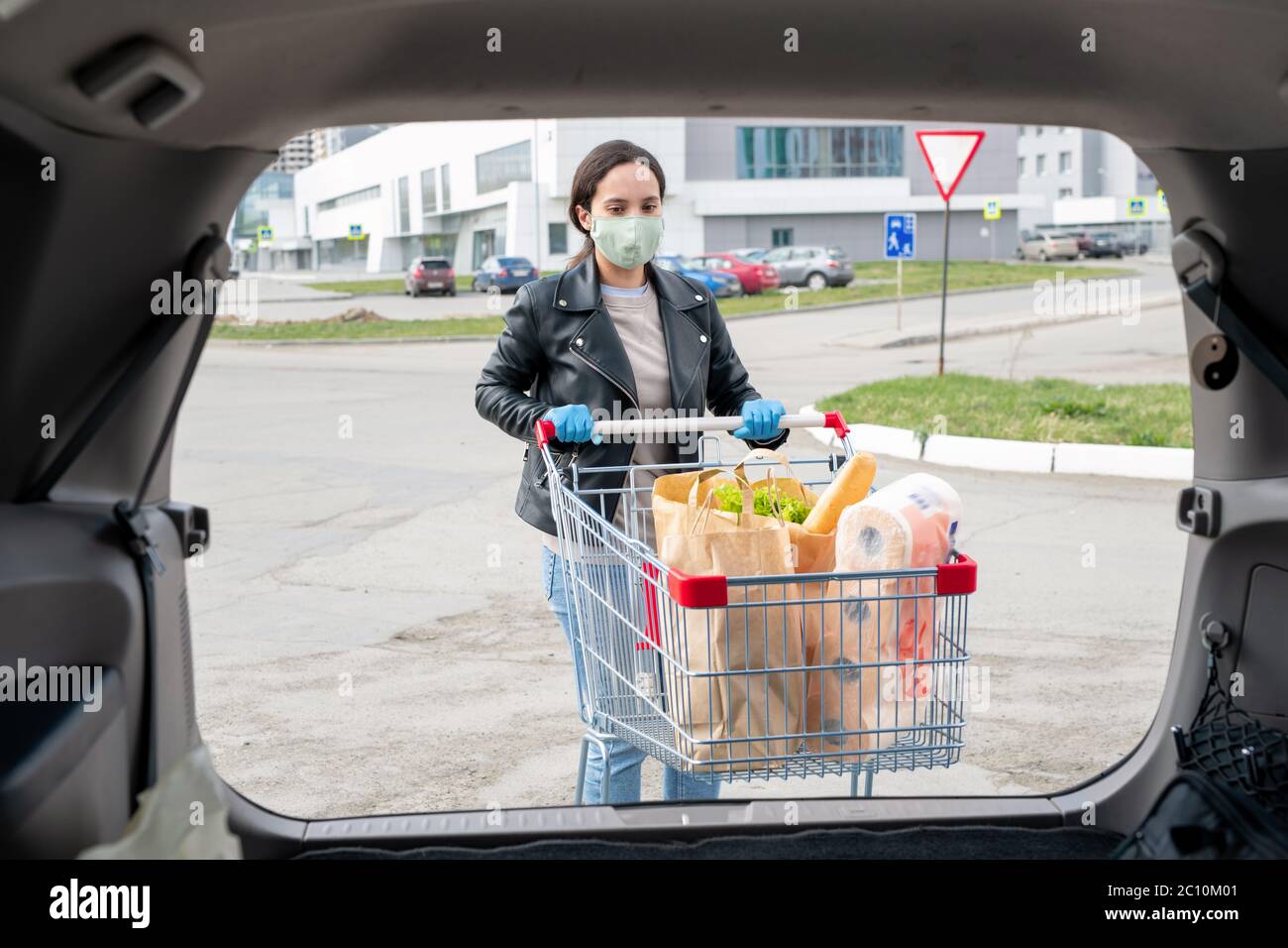 Young woman in cloth mask and gloves pushing shopping cart to car to ...