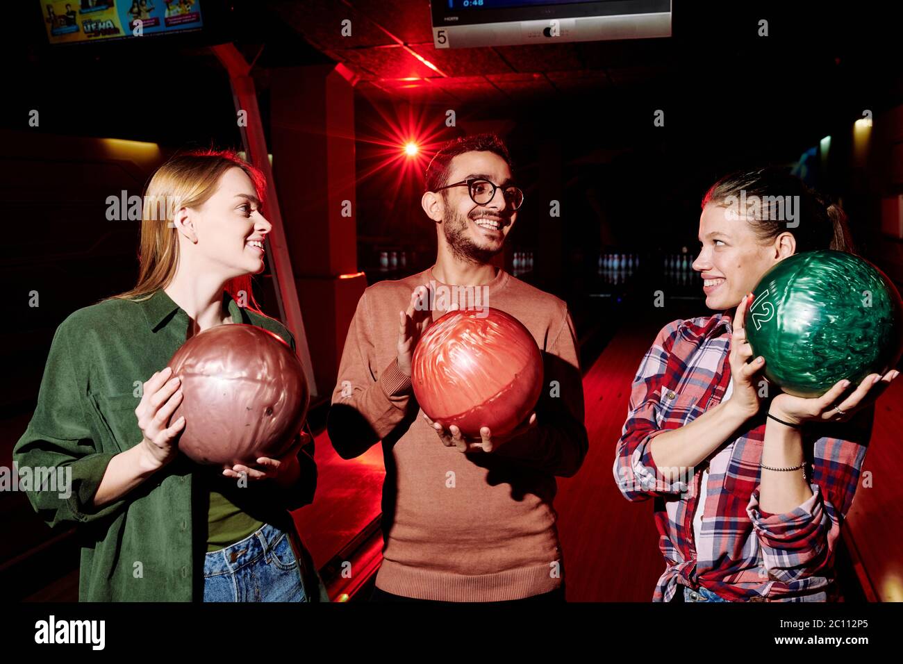 Happy young intercultural man with bowling ball talking to pretty girls ...