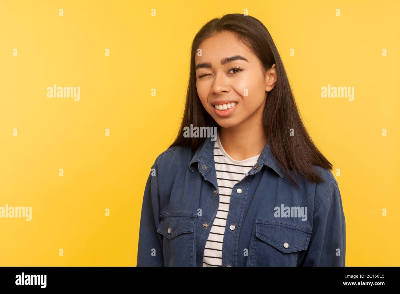 Portrait of lovely positive friendly girl in denim shirt standing, winking playfully and smiling ...