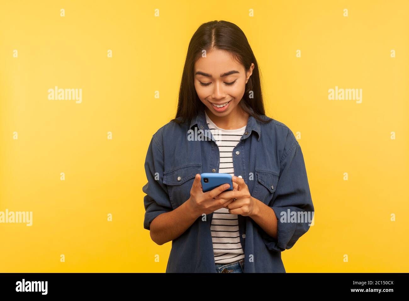 Portrait of happy girl in stylish denim shirt typing message and smiling, dialing number calling ...