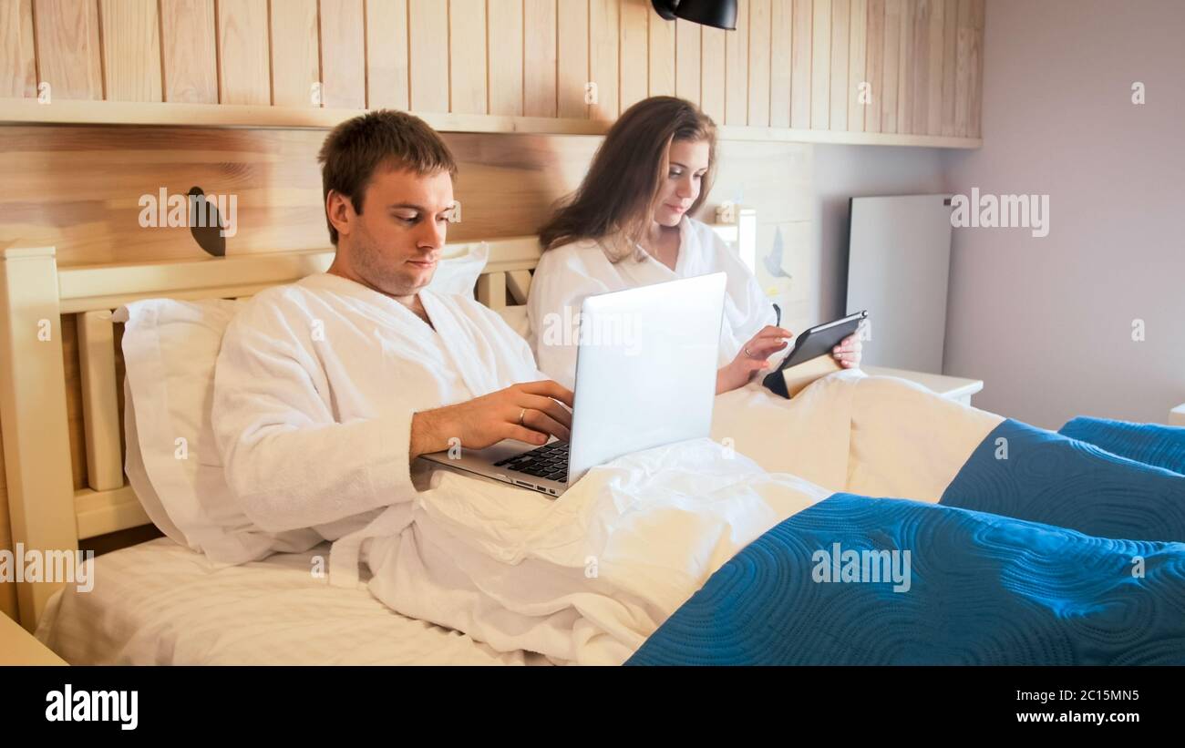 Young busy couple lying in bed at morning and working on laptop and ...