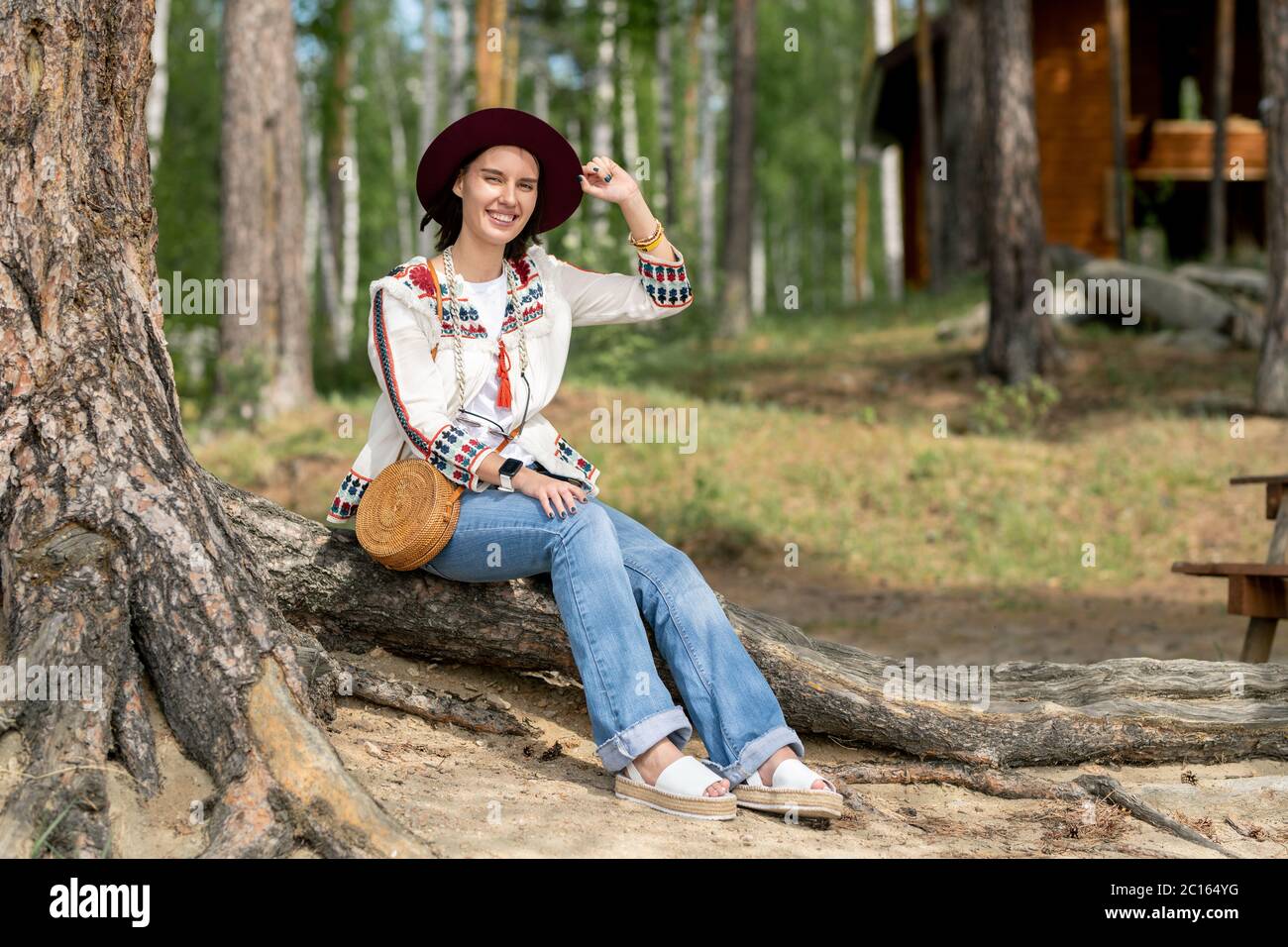 Portrait of smiling pretty young woman in jeans and hat sitting on root ...