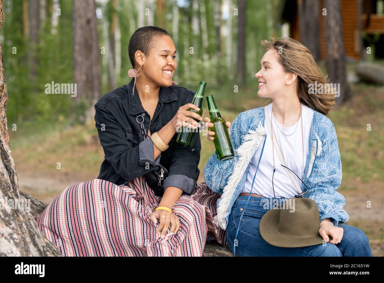 Positive multi-ethnic girls sitting in forest and clinking bottles ...
