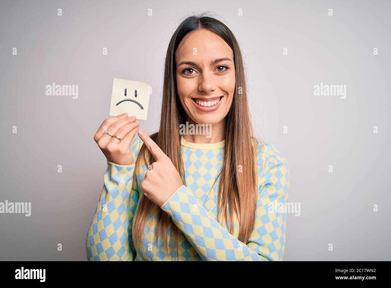 Young blonde woman holding sticky paper note with sad face emoticon ...