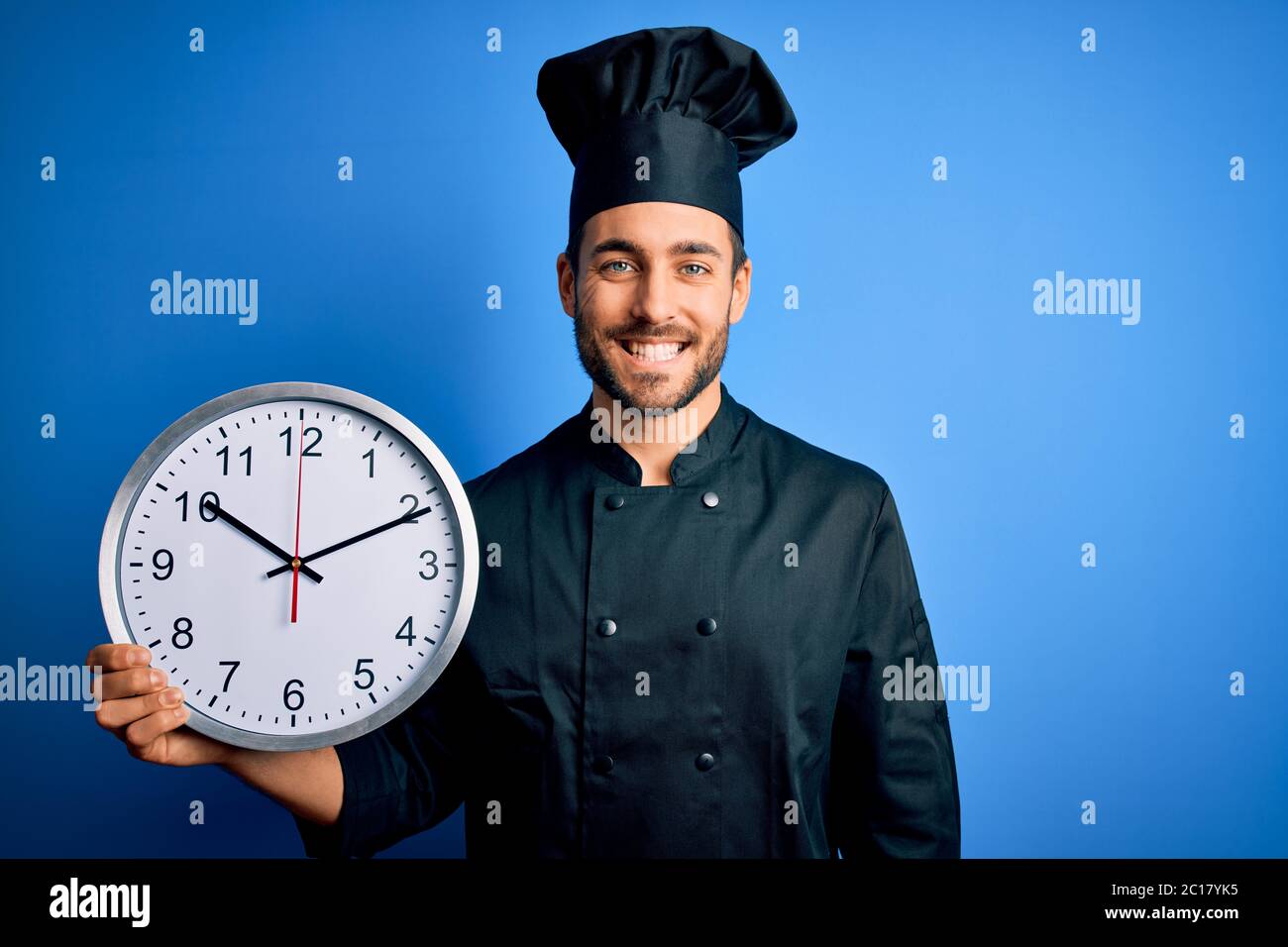 Young handsome cooker man with beard wearing uniform holding clock ...