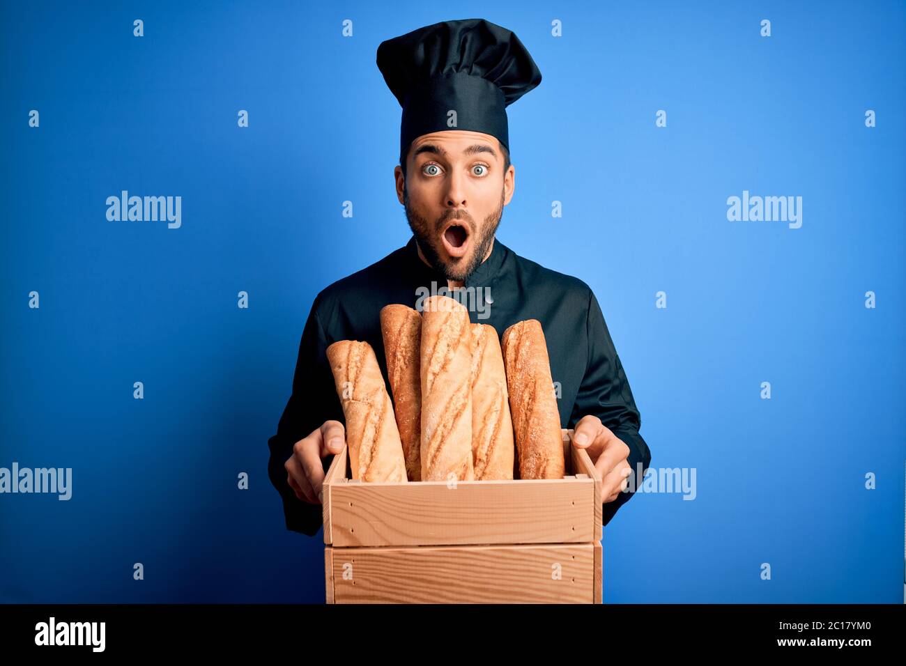 Young cooker man with beard wearing uniform holding box with bread over ...