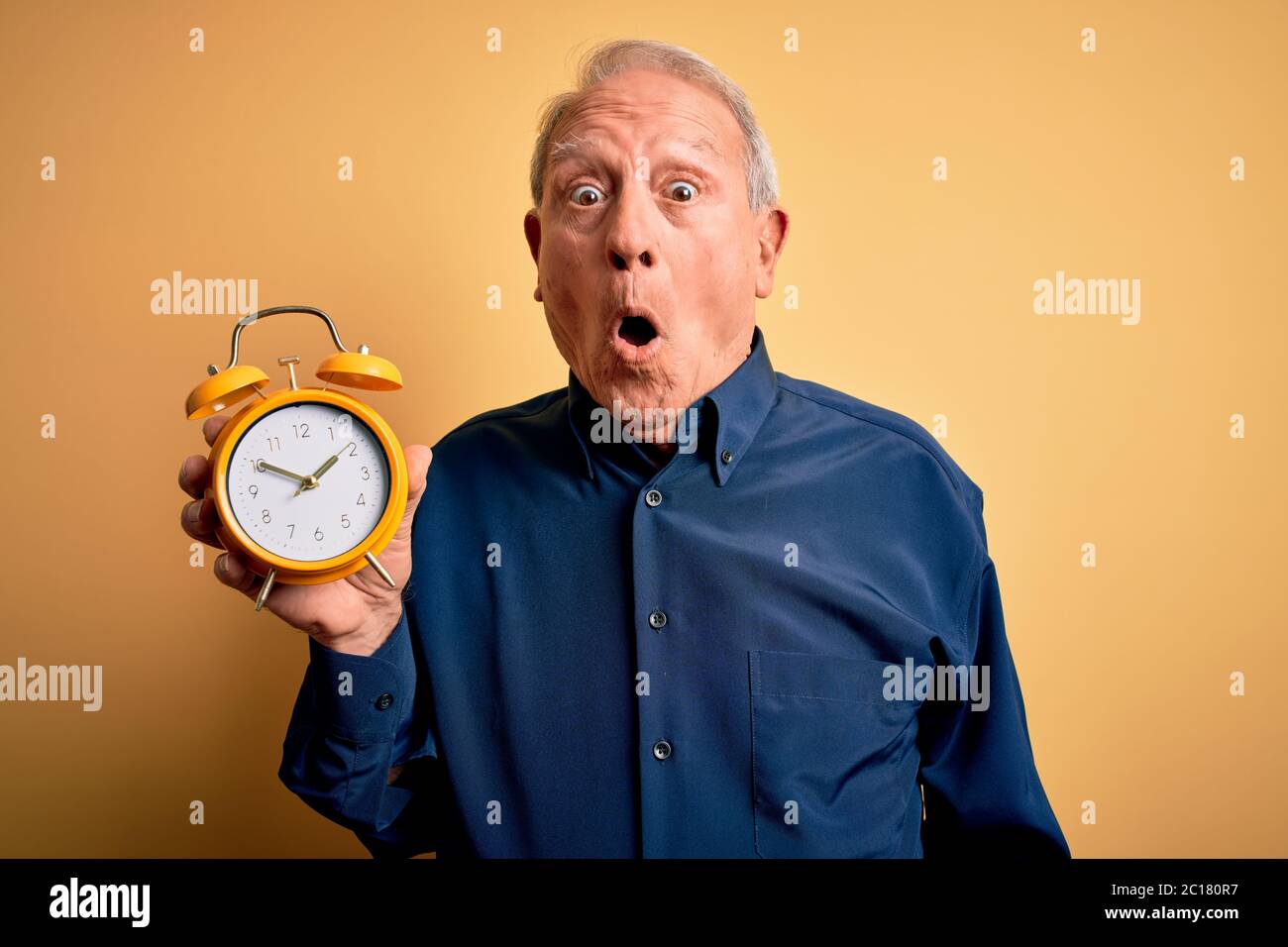 Senior grey haired man holding vintage alarm clock over yellow ...