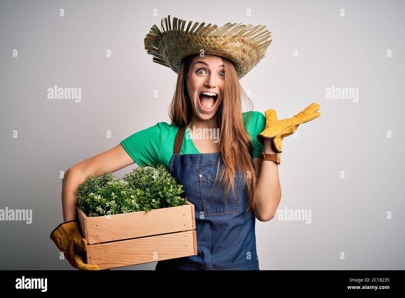 Young beautiful redhead farmer woman wearing apron and hat holding box ...