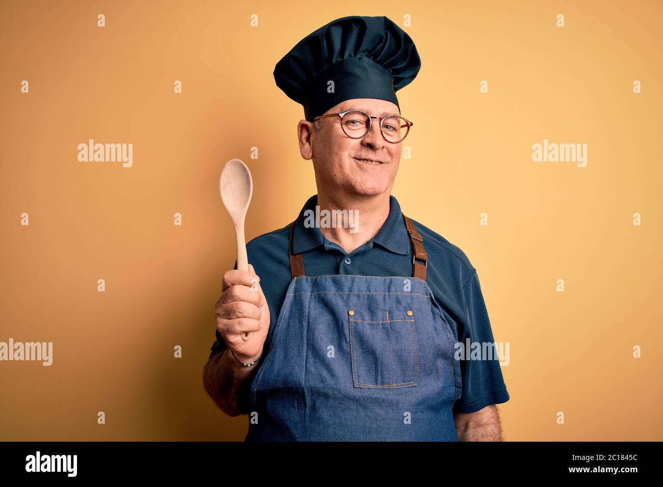 Middle age cooker man wearing apron and hat holding wooden spoon over ...