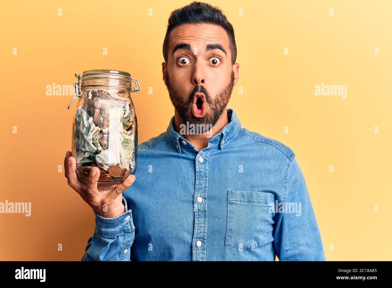 Young hispanic man holding jar with savings scared and amazed with open ...