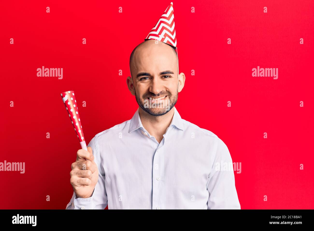 Young handsome bald man wearing party hat and trumpet looking positive ...