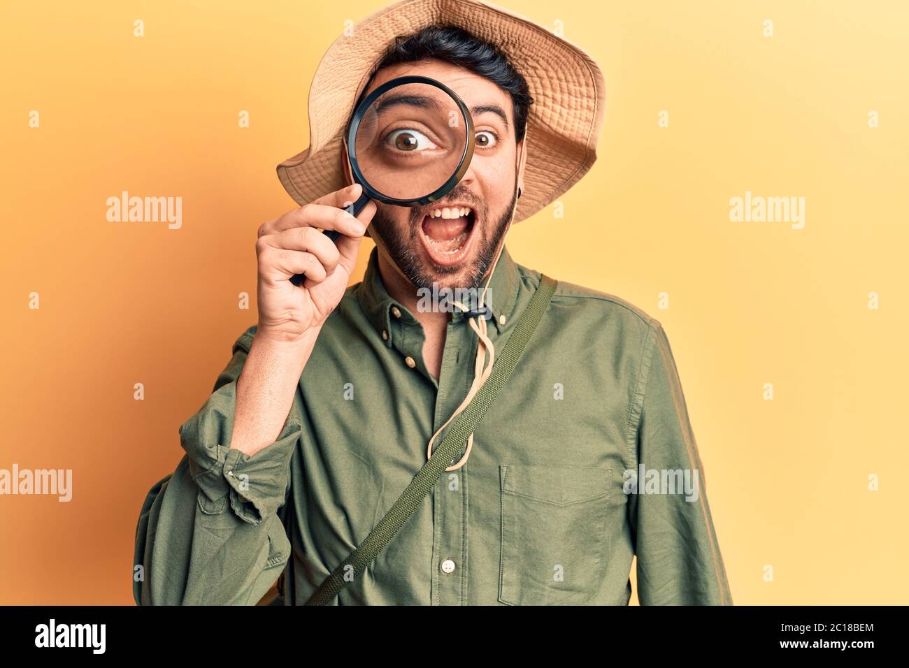 Young hispanic man wearing explorer hat holding magnifying glass scared ...