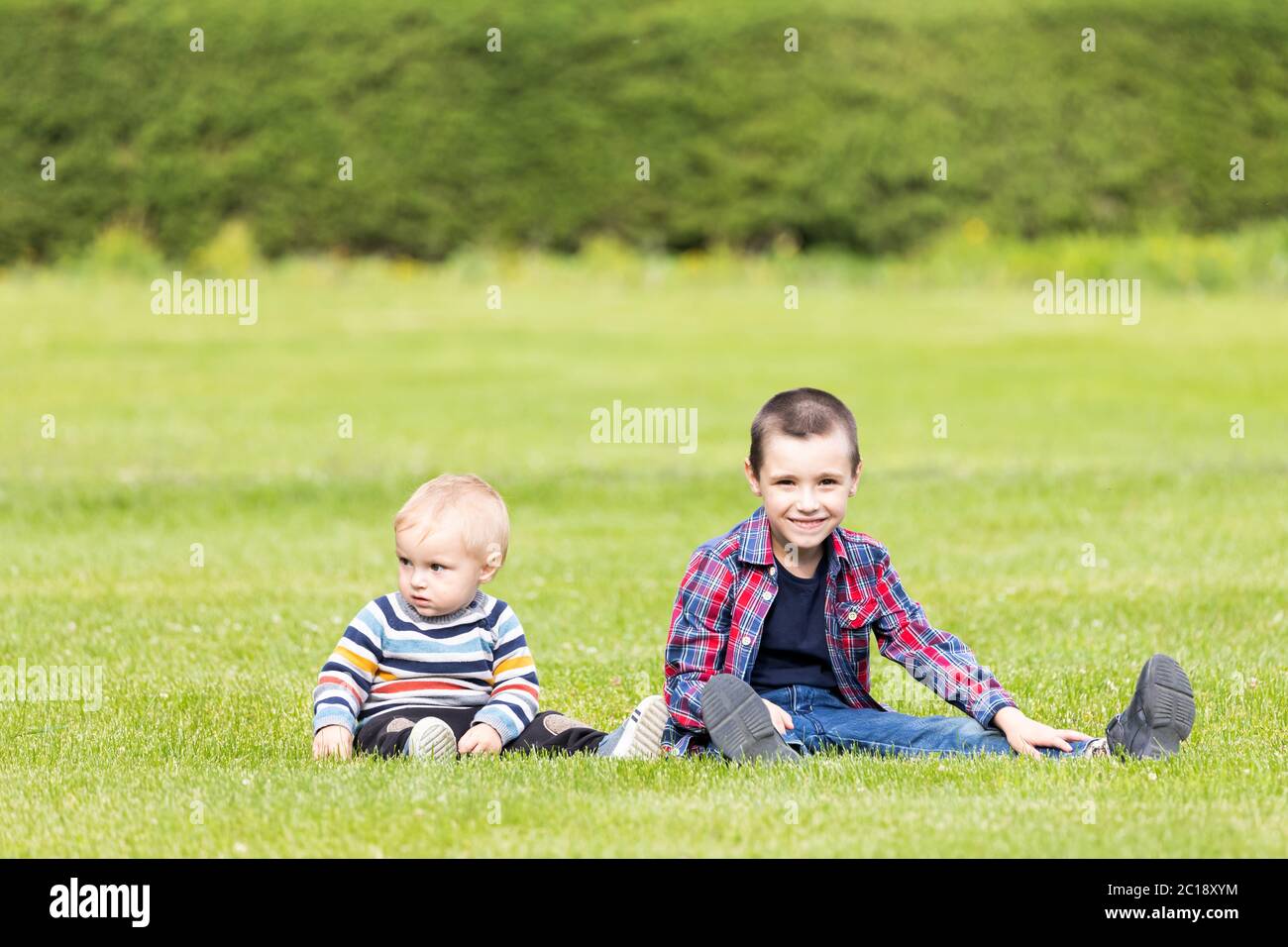 Two cheerful boy brothers of different ages play fun, sit on the grass ...