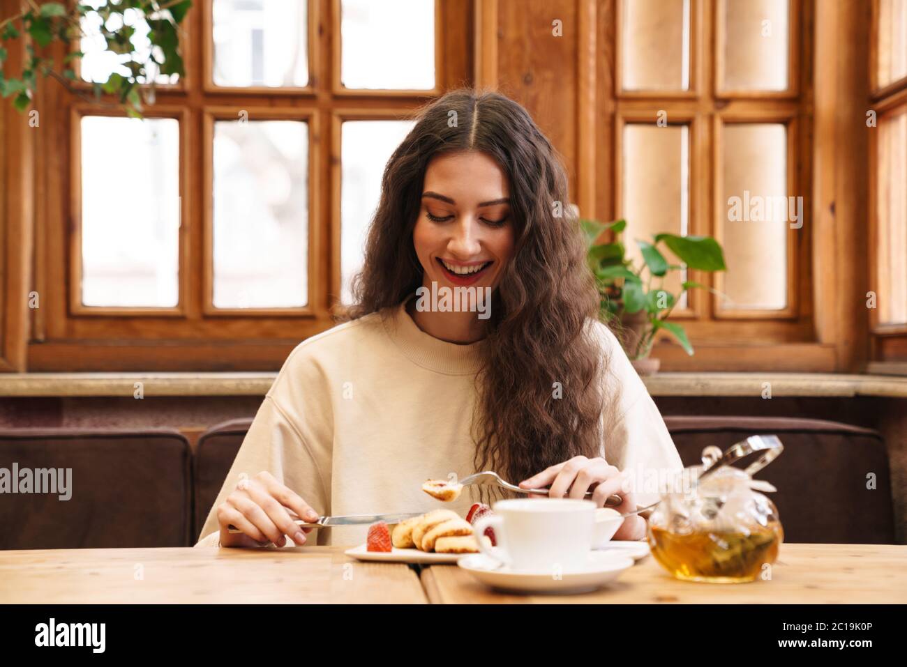 Image of happy beautiful young woman smiling and eating pancakes while ...