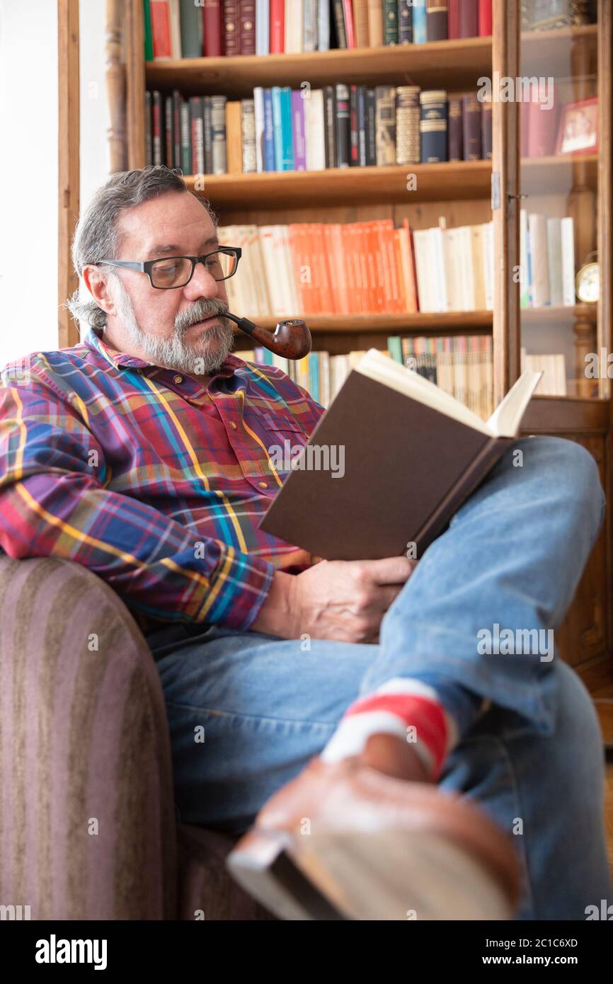 Portrait of older man reading a book next to his own library while ...