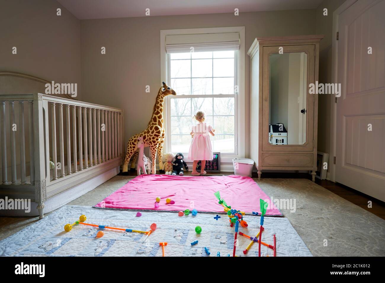 Toddler girl looks out the window of her nursery, toys on the floor ...