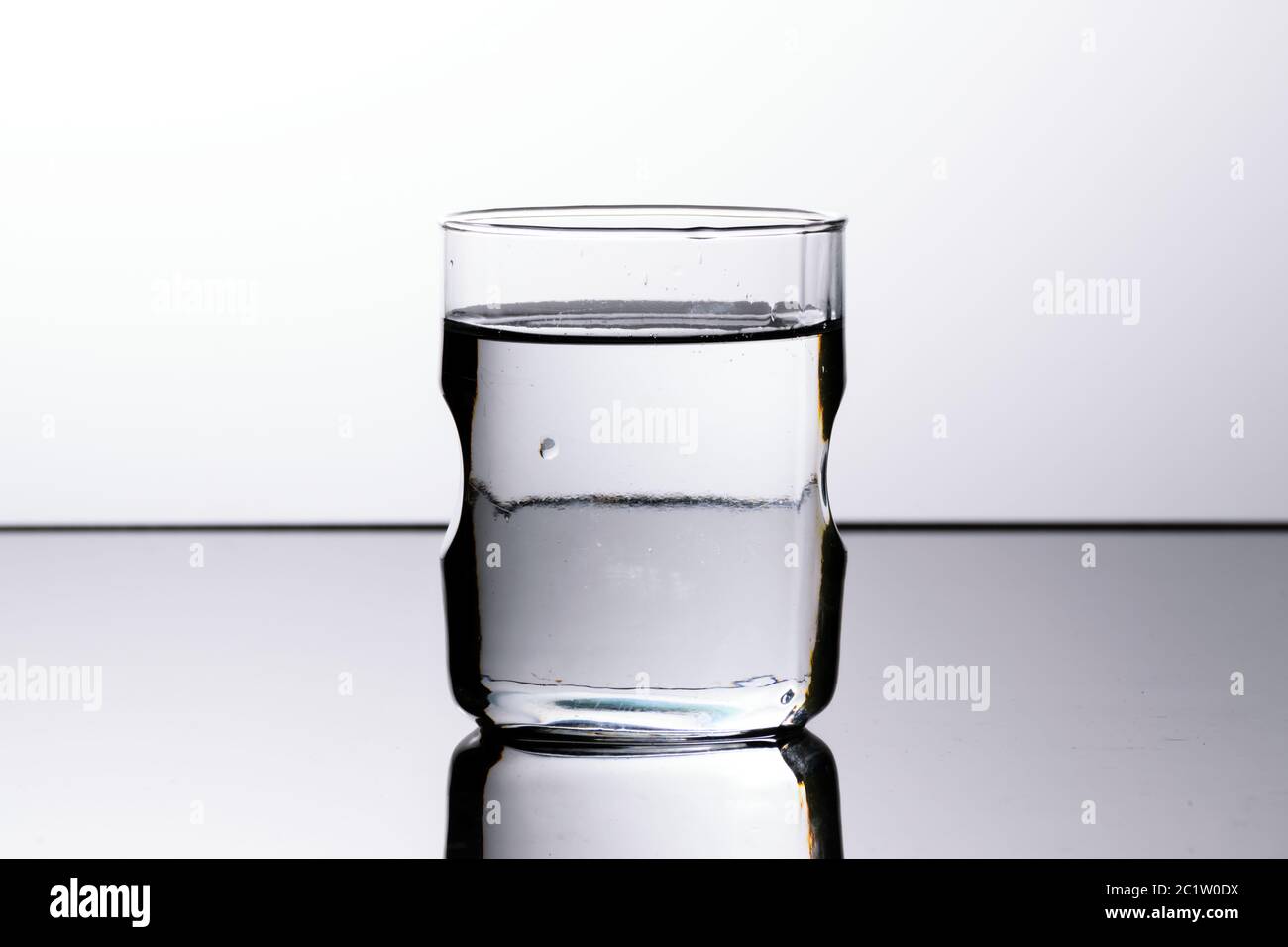 A glass of transparent water kept on a reflective surface in a white ...