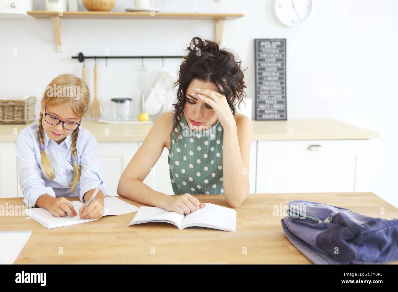 Сute school girl wearing eyeglasses doing homework with mother at home ...