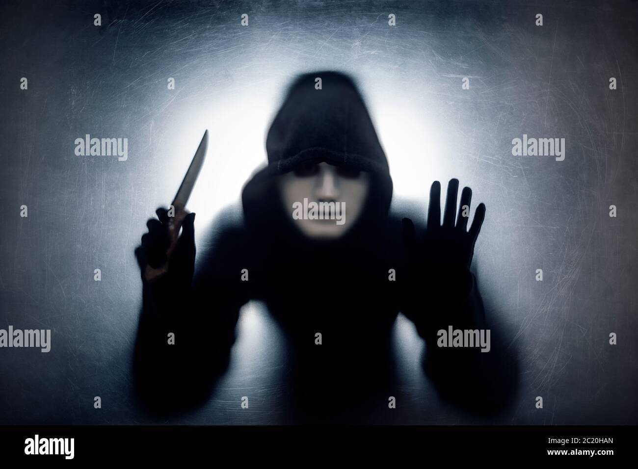 Man in a hood and white mask with knife behind a dusty scratched glass ...