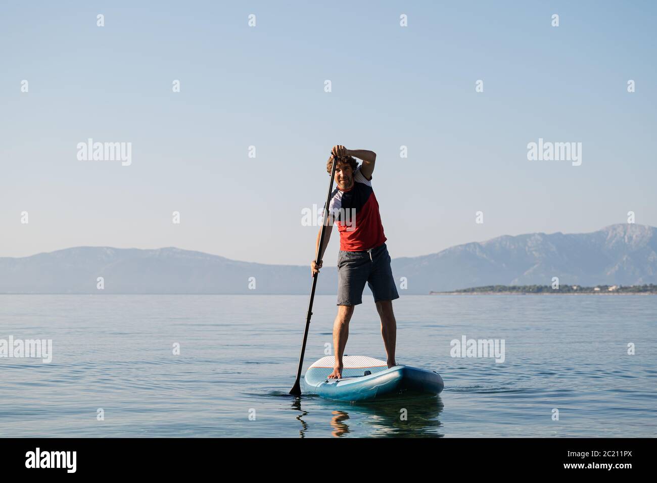 Fit young man standing on sup board paddling on calm morning sea water ...