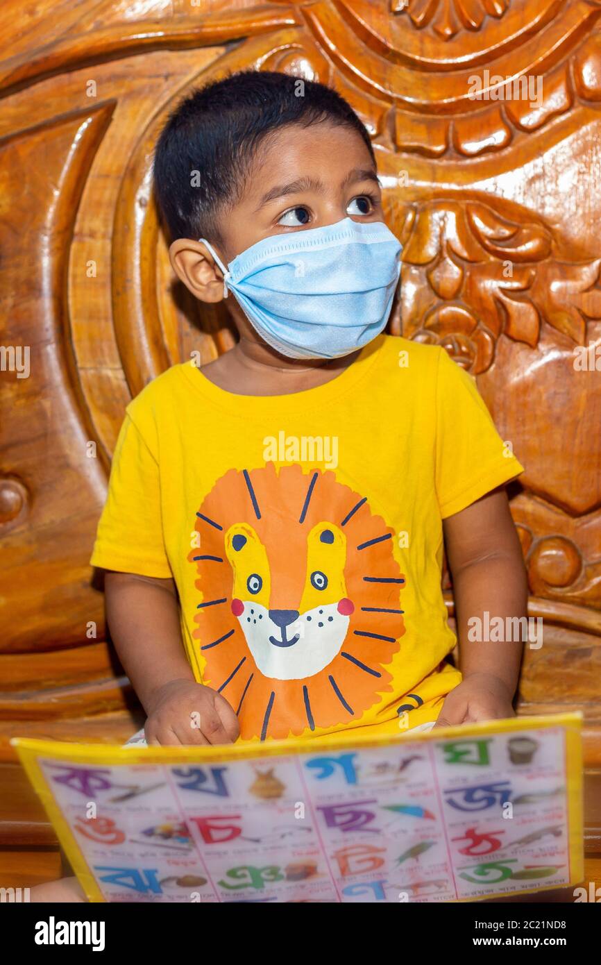 A baby boy is wearing a face mask and sitting on a bed reading a book ...