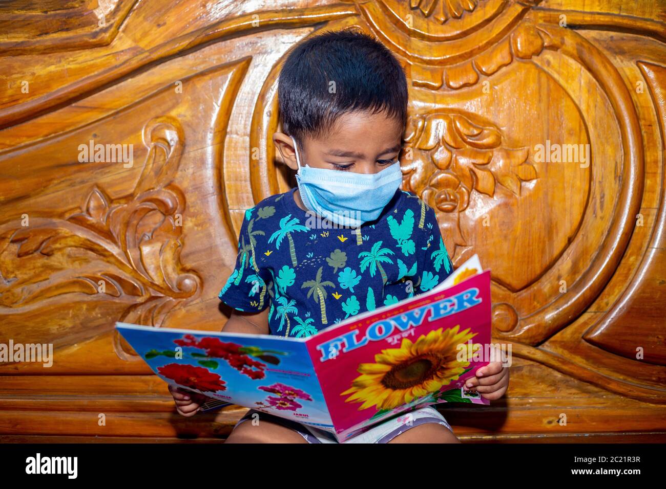 A baby boy is wearing a face mask and sitting on a bed reading a book ...