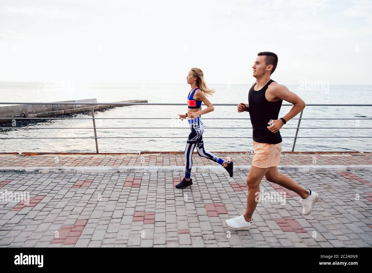 Side view of attractvive young couple in sportswear, jogging on quay ...