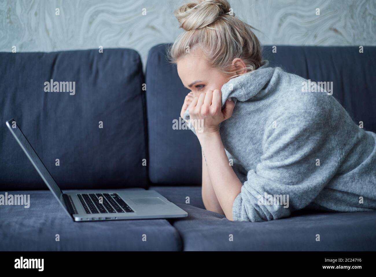 Scaried blond woman lying prone on sofa and looking on laptop computer ...