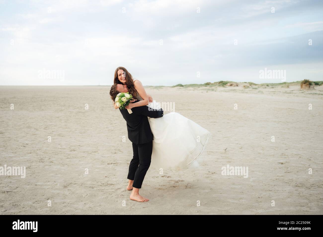 Wedding couple standing and hugging at sandy beach. Groom lifting bride to air. Smiling cheerful ...