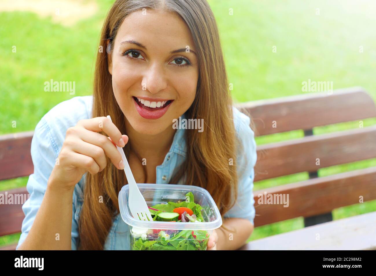 Happy smiling woman eating salad sitting in the park looks at camera ...
