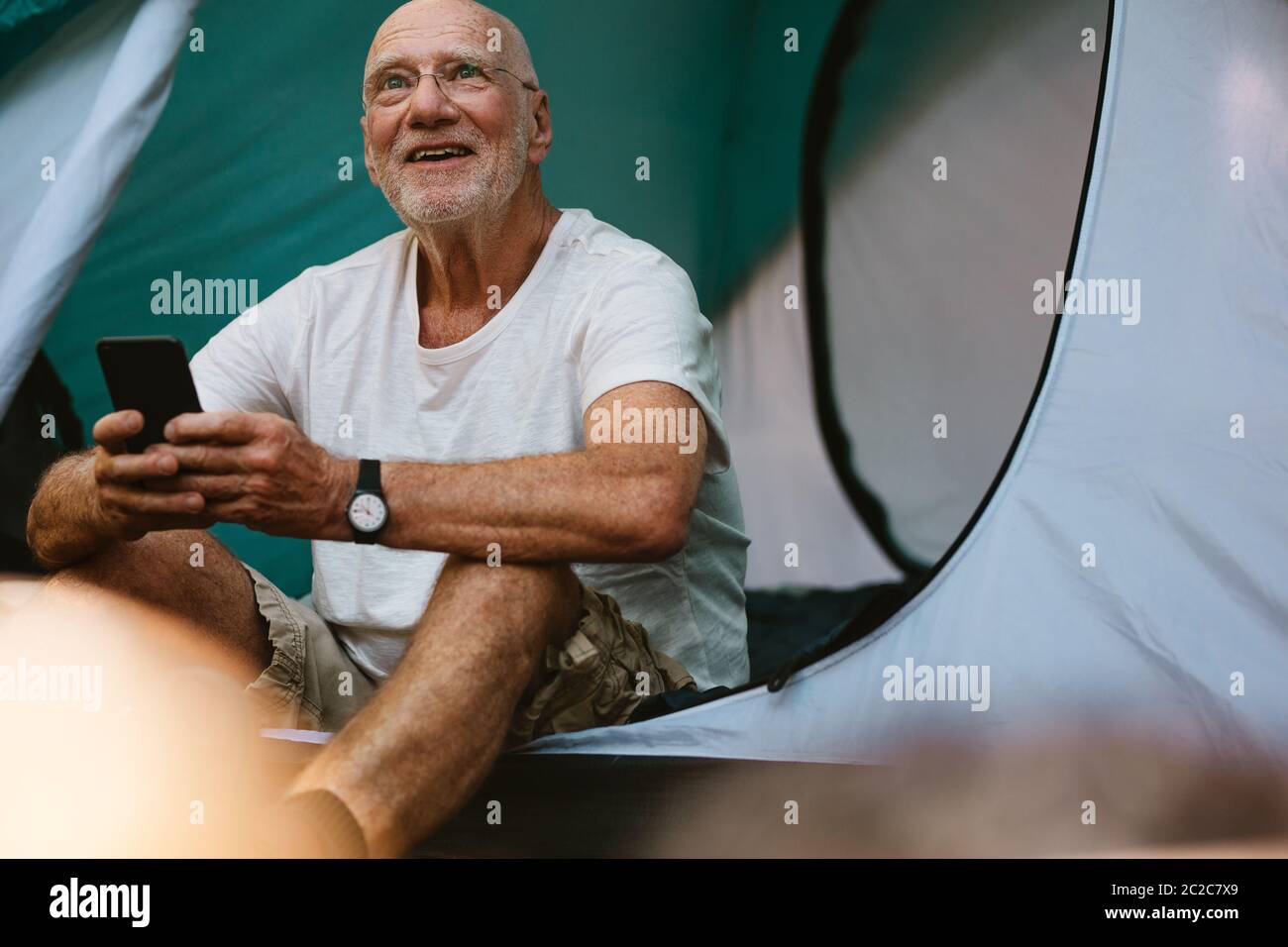 Smiling senior man sitting in tent with his mobile phone. Retired man ...